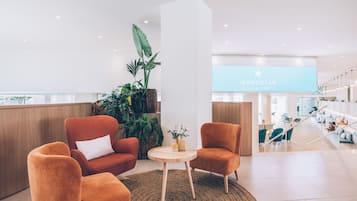 Um saguão de hotel moderno com balcão de recepção, assentos espaçosos em tom de laranja, uma grande planta em vaso e a placa do hotel à vista.