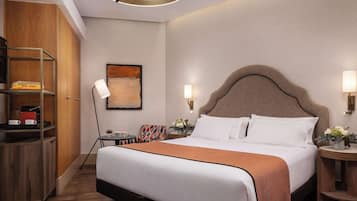 Quarto de hotel com cama grande, guarda-roupa de madeira, escrivaninha com abajur e uma obra de arte na parede.