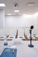 Sala de conferências com fileiras de cadeiras brancas, projetor fixado no teto, microfone em pedestal e mesa com garrafa de água, copo e caderno.