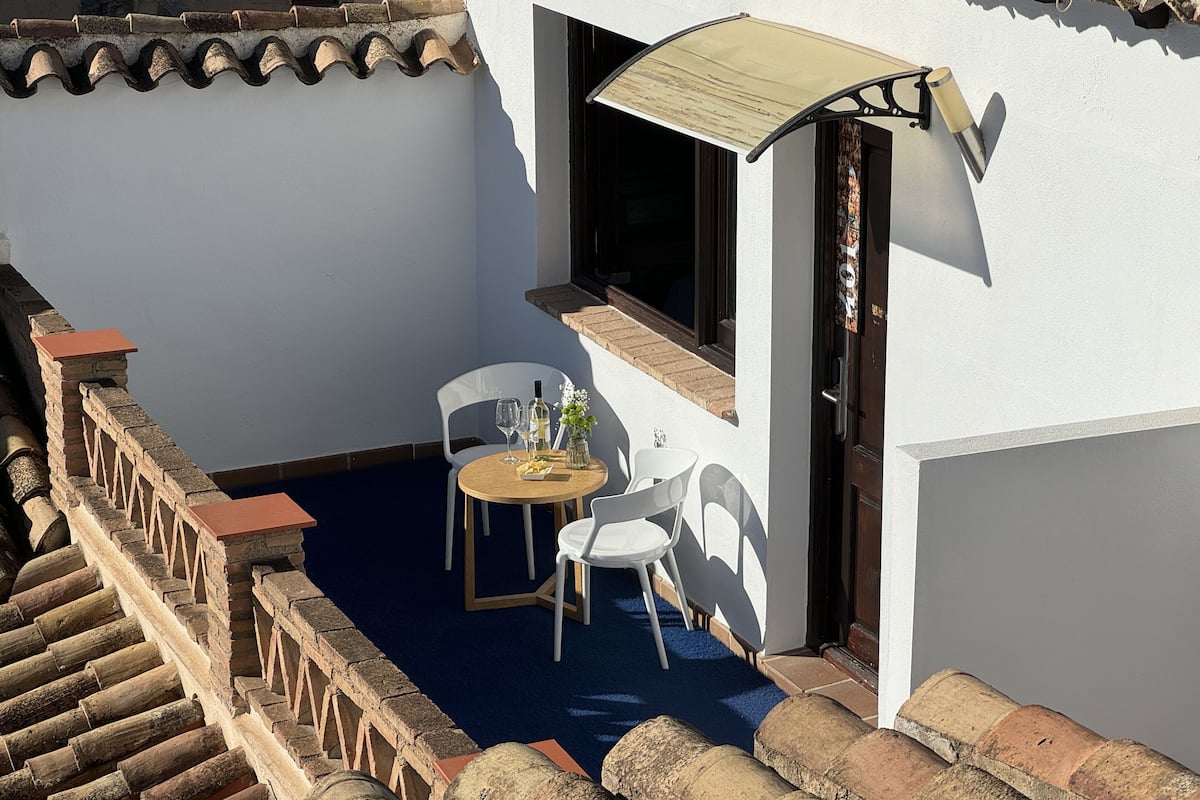 Um pequeno pátio exterior com uma mesa redonda e duas cadeiras brancas, uma janela com toldo bege e um parapeito de pedra.