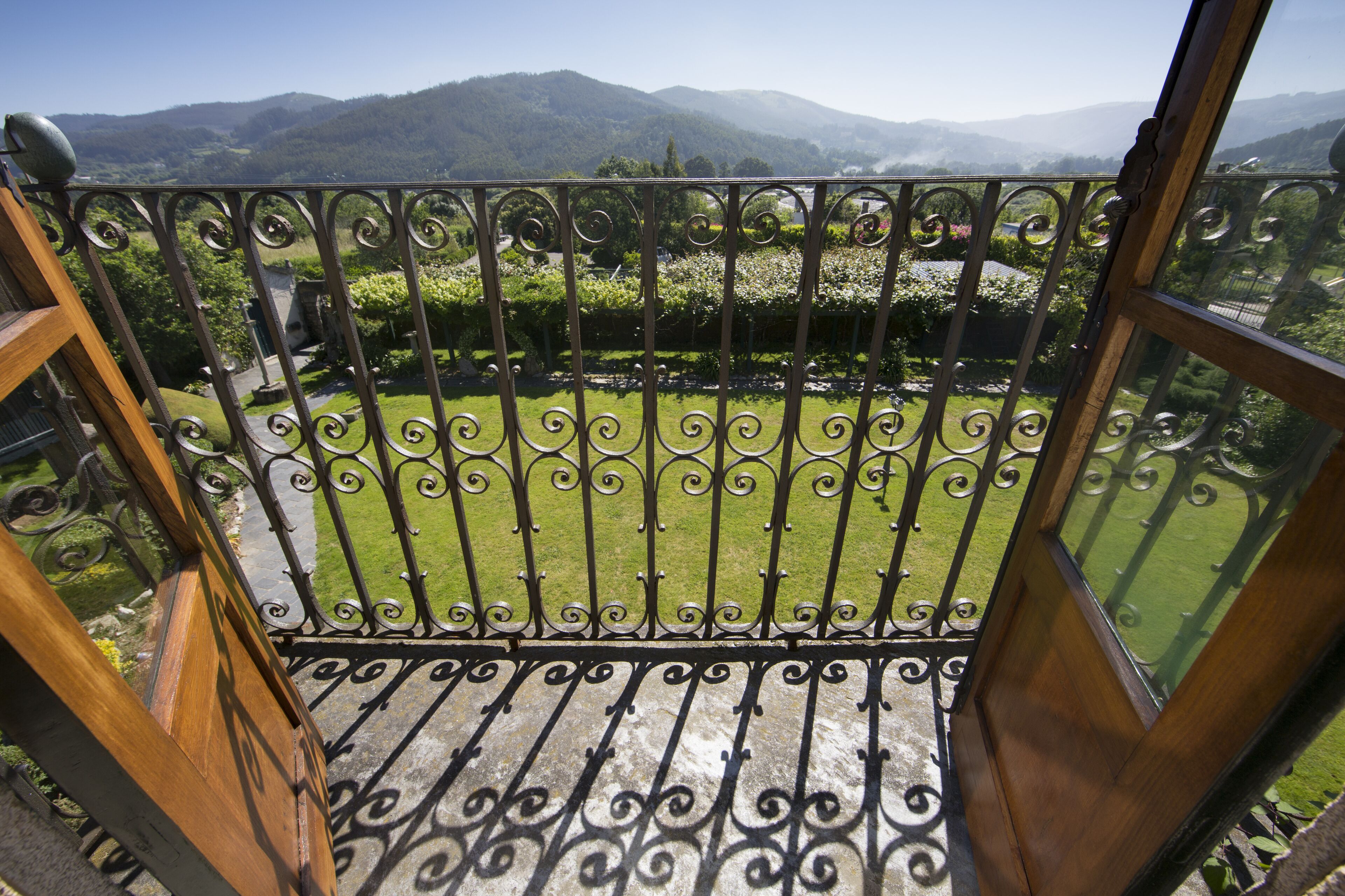 Varanda com grades de ferro forjado, vista para um jardim exuberante e montanhas ao longe.