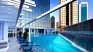 Uma piscina no terraço com guarda-corpos de vidro, áreas de estar e vista para edifícios ao redor.