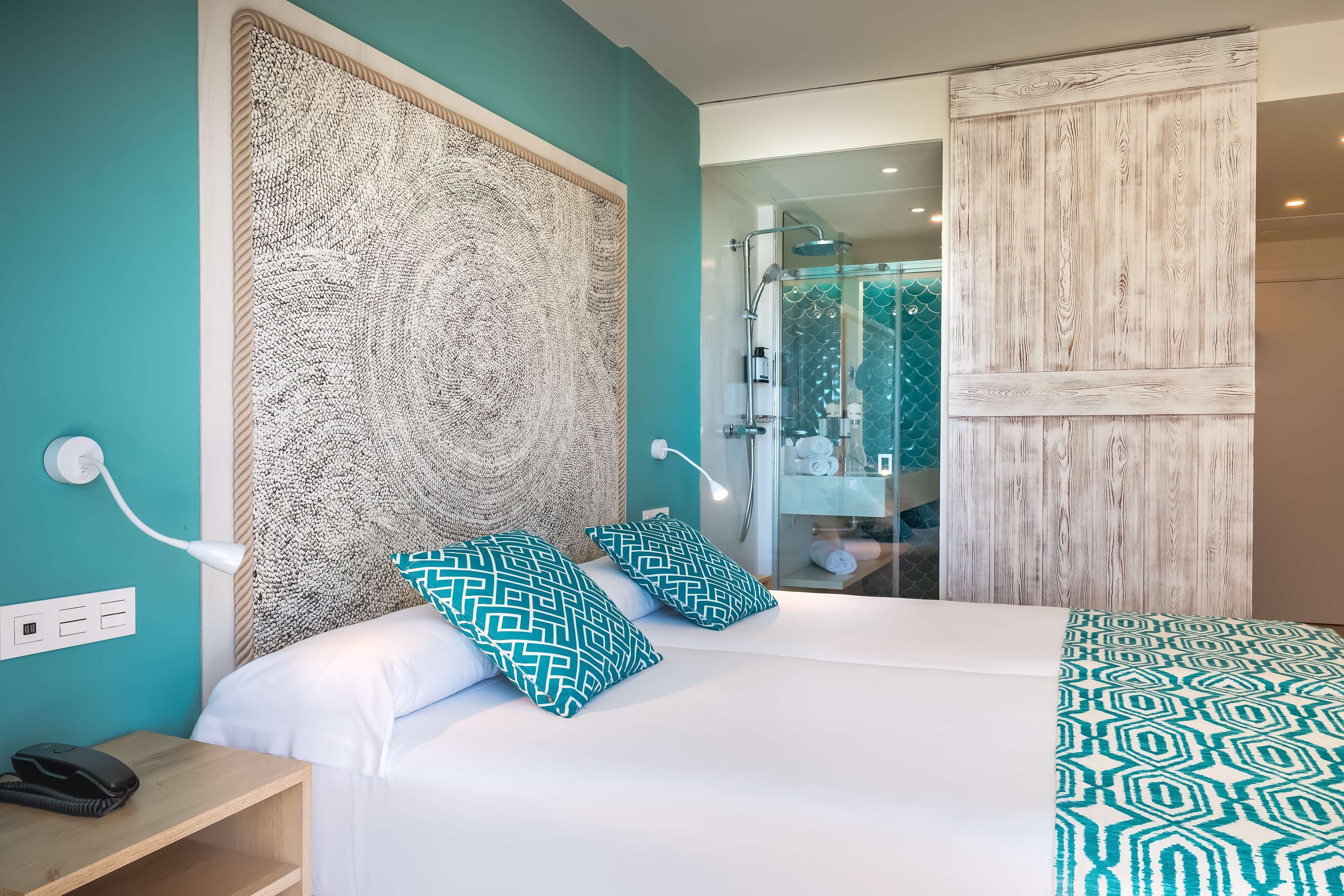 Quarto de hotel com uma cama grande, duas almofadas estampadas, mesa de cabeceira com telefone e cabeceira fixada na parede.