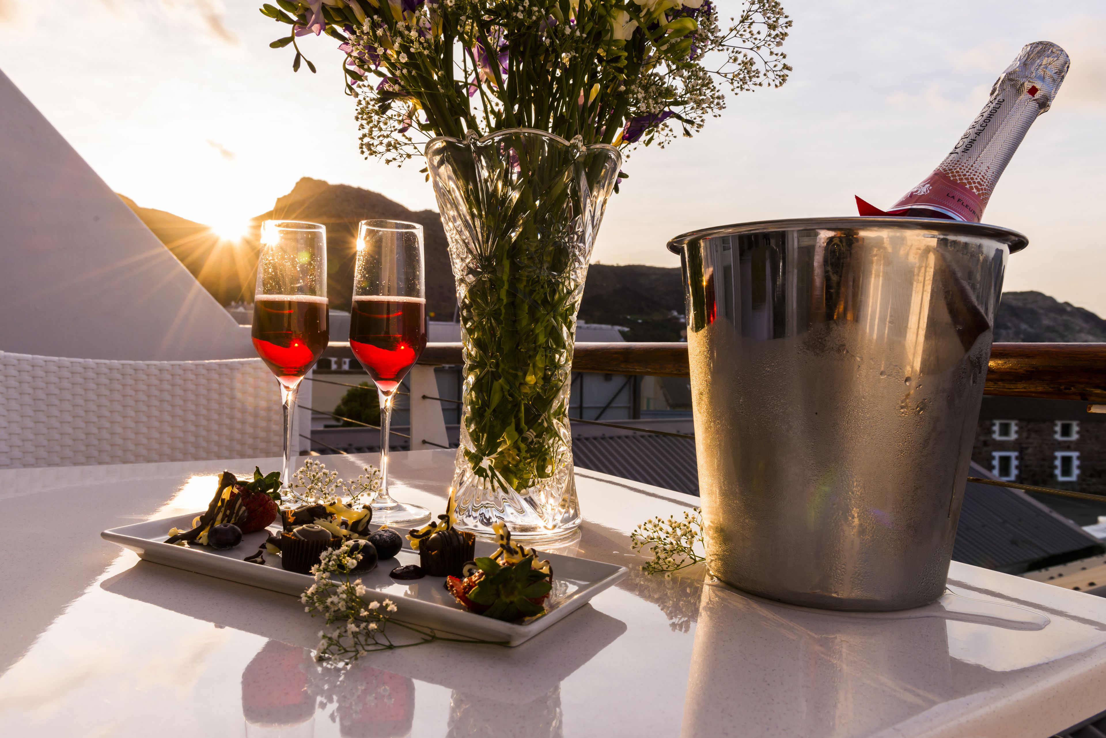 Uma mesa com uma garrafa de champanhe, duas taças de vinho tinto, um vaso de flores e um prato de comida.