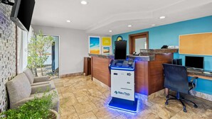 Uma área de recepção com um quiosque NexTap de autoatendimento, um balcão de recepção, um computador e um espaço de estar com uma planta.