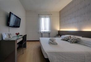 Quarto de hotel com cama, escrivaninha, televisão e janela com cortinas.
