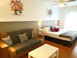Quarto de hotel moderno com um sofá cinzento, uma mesa de centro branca, uma cama com várias almofadas e duas luminárias fixadas na parede.