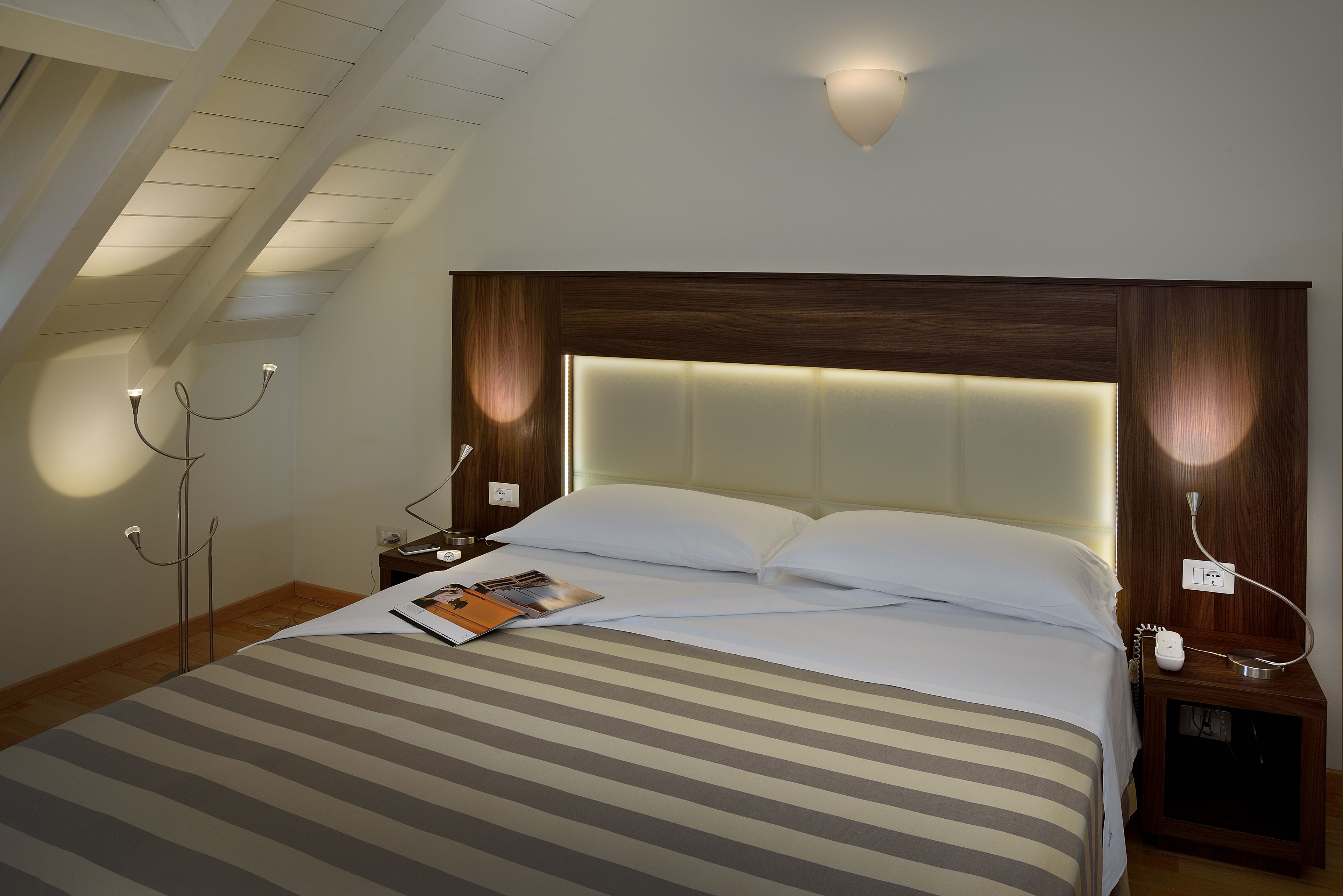 Quarto de hotel com cama grande, cabeceira de madeira, luminárias de cabeceira e uma revista sobre a cama.