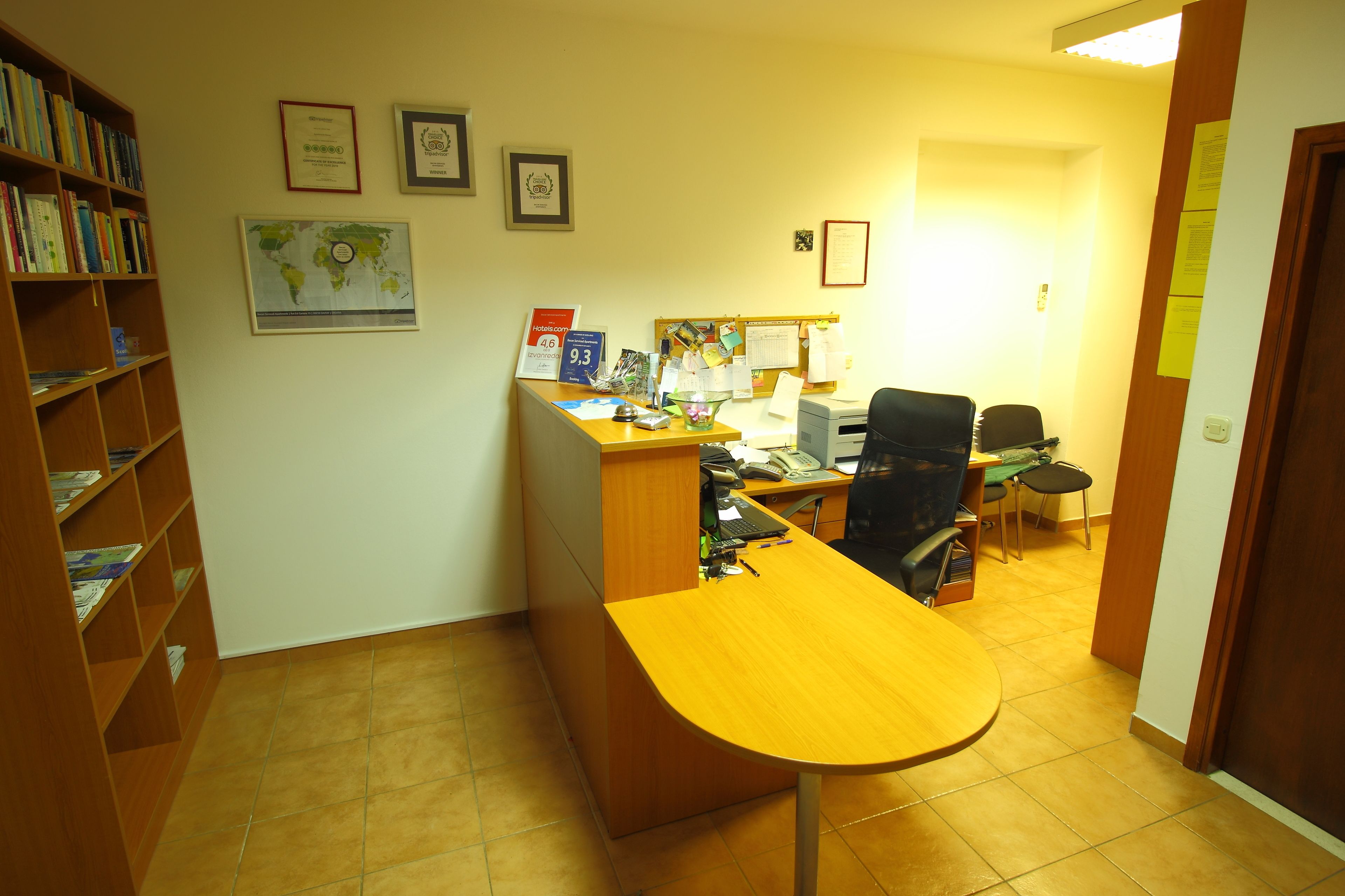 Um escritório com uma mesa de madeira, um computador e uma estante. Há certificados emoldurados na parede.