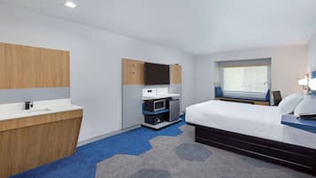 Quarto de hotel com cama, escrivaninha com computador, micro-ondas e pequena cozinha.