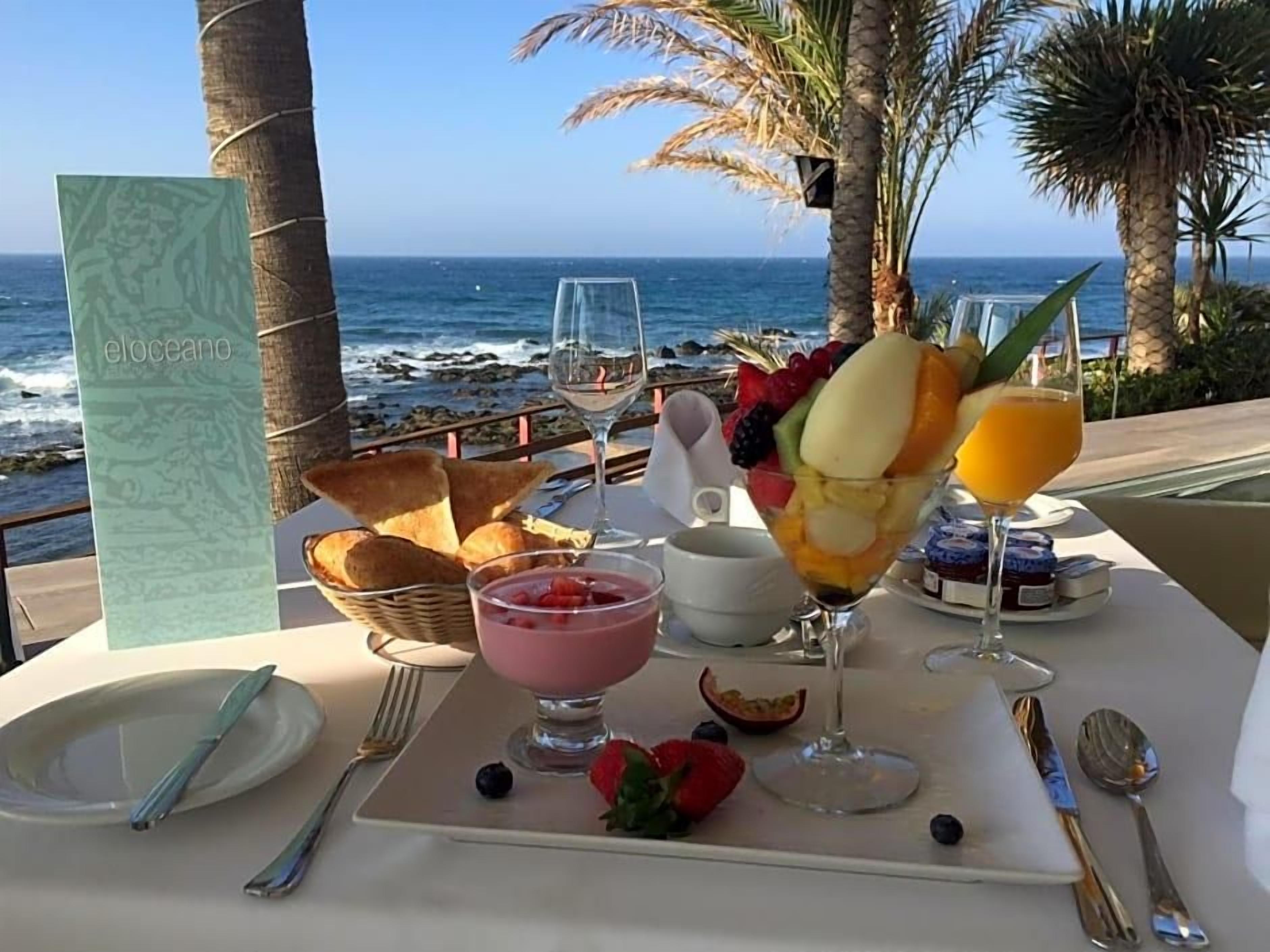 Mesa posta com copo de suco de laranja, tigela de frutas, copo de água, prato com faca e colher, e cardápio intitulado 'eloceano', em uma praia com palmeiras e o oceano ao fundo.