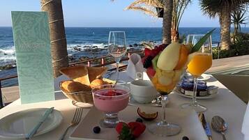 Mesa posta com copo de suco de laranja, tigela de frutas, copo de água, prato com faca e colher, e cardápio intitulado 'eloceano', em uma praia com palmeiras e o oceano ao fundo.