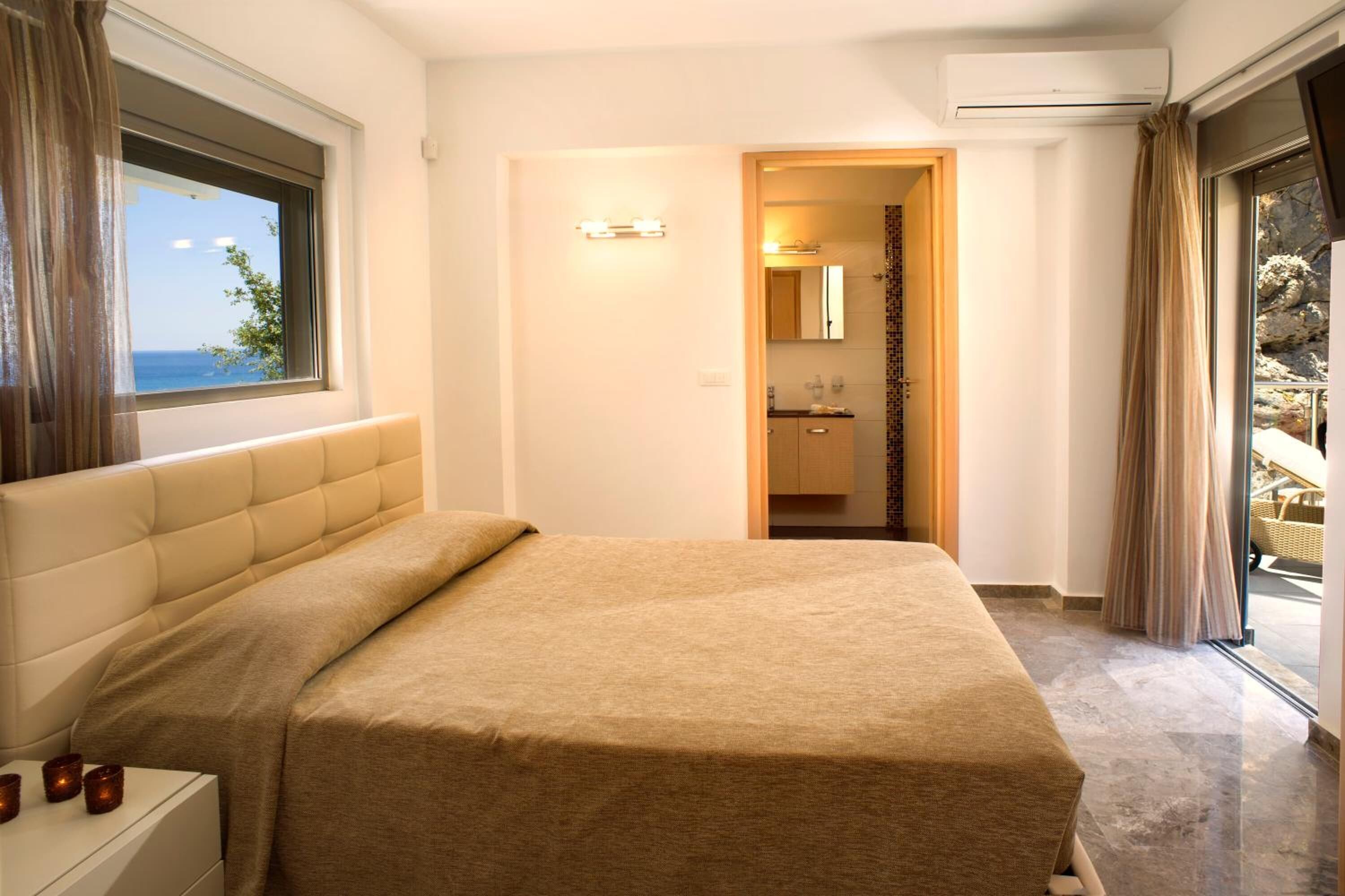 Quarto de hotel com cama, mesa de cabeceira, janela com vista, banheiro com espelho e porta de vidro deslizante.