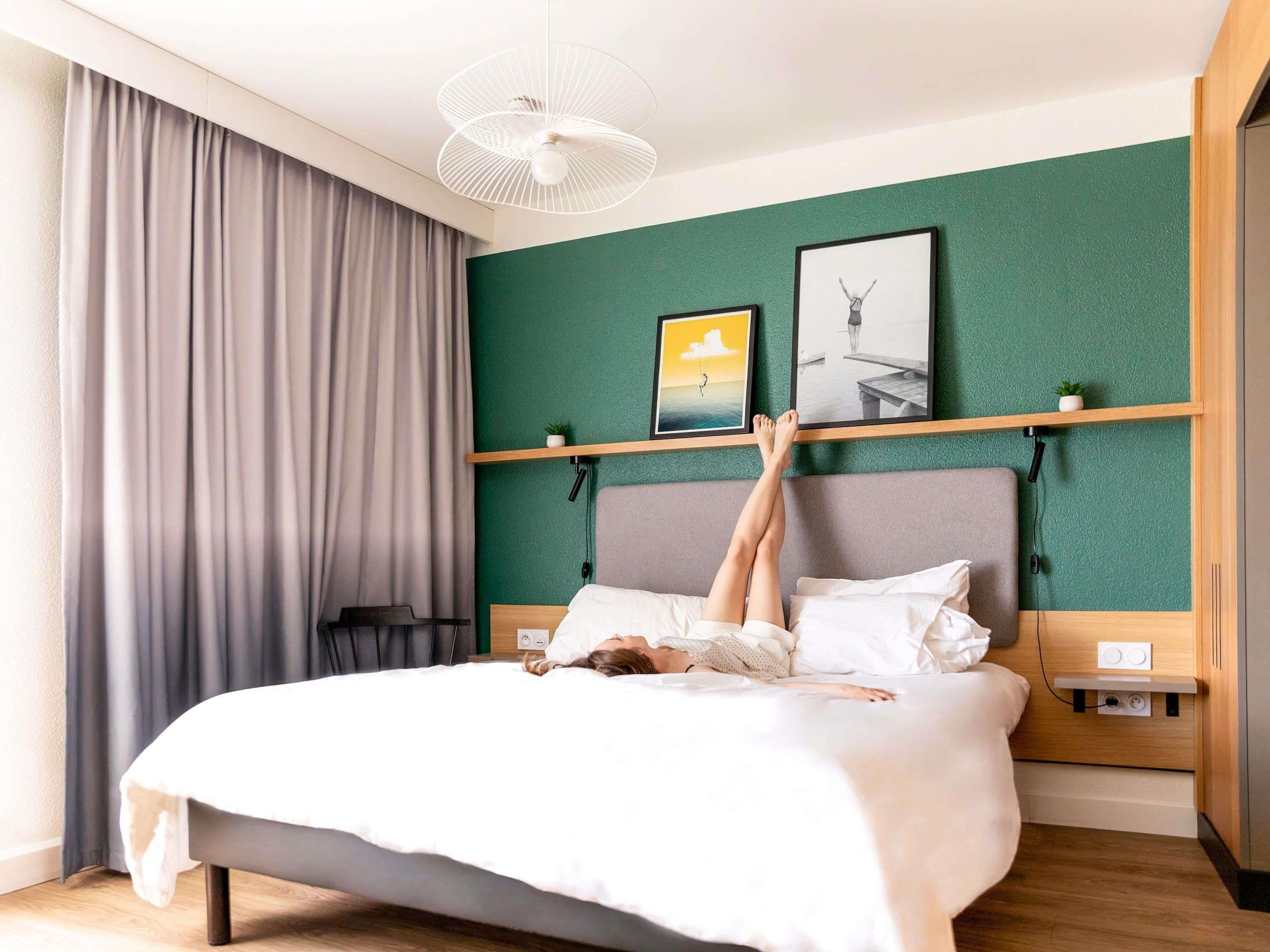 Quarto de hotel moderno com cama, cabeceira, prateleira fixada na parede com obras de arte, ventilador de teto e janela com cortinas.