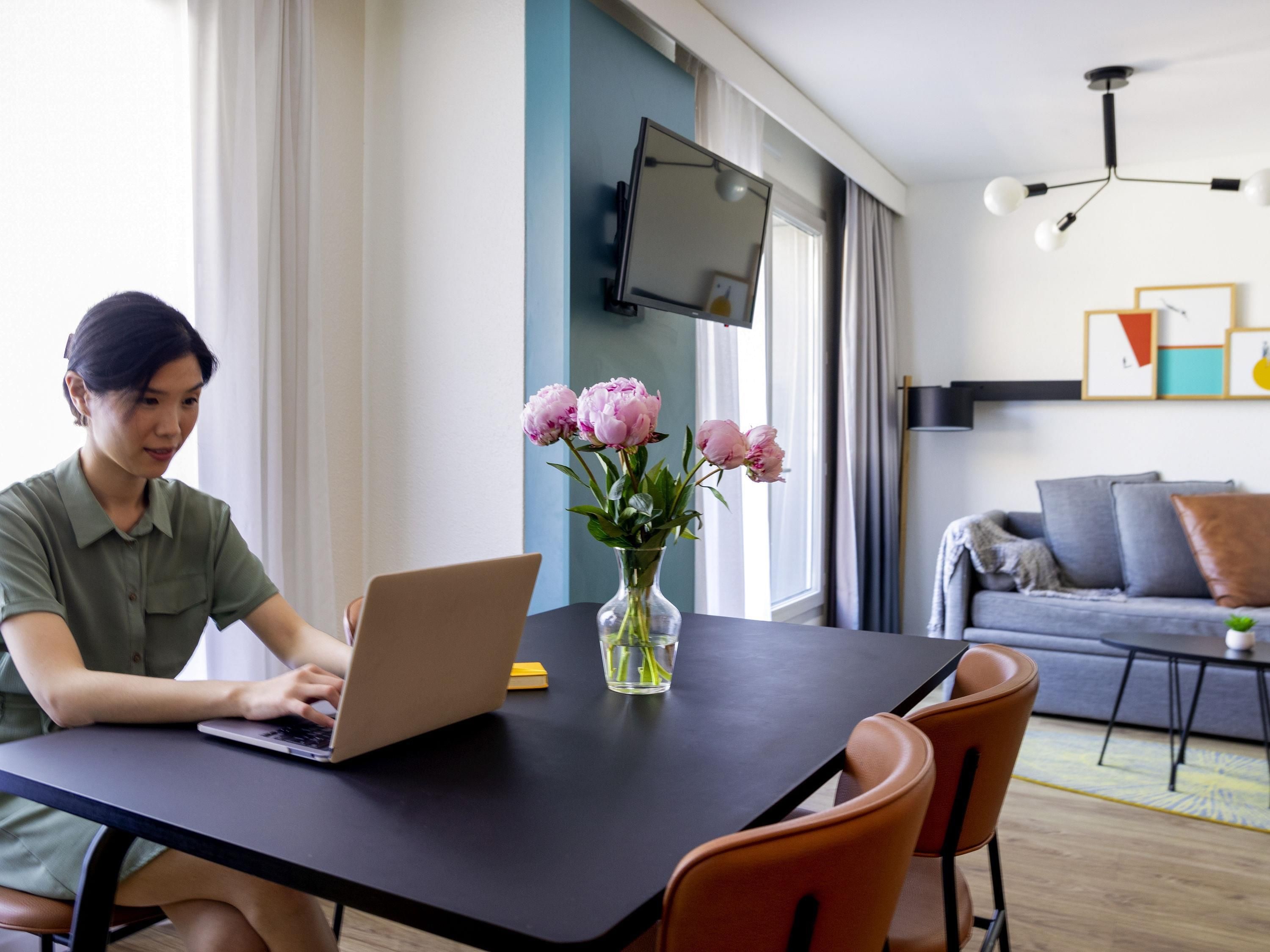 Uma pessoa trabalhando em uma mesa com um laptop, um vaso de flores e uma televisão na parede.