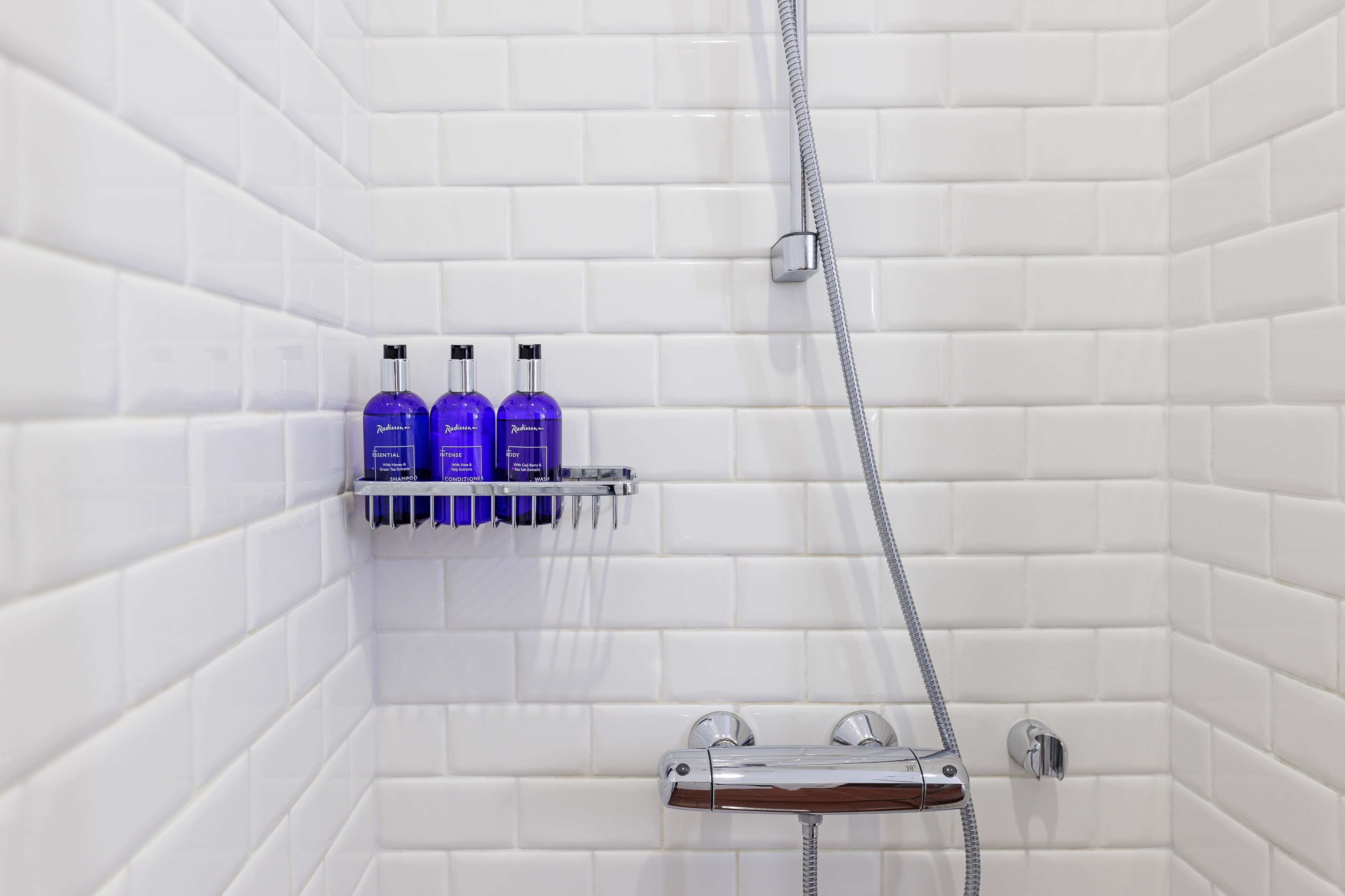 Um banheiro com paredes revestidas de azulejos brancos, um chuveiro com haste móvel e uma prateleira com três frascos de shampoo.