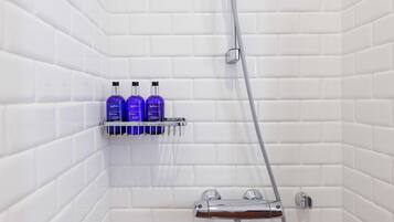 Um banheiro com paredes revestidas de azulejos brancos, um chuveiro com haste móvel e uma prateleira com três frascos de shampoo.