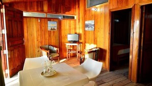 Interior de uma cabana de madeira com uma mesa de jantar, cadeiras, uma televisão e quadros nas paredes.