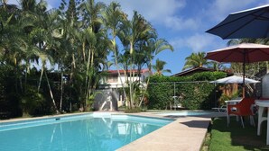 Piscina com borda azulejada, cercada por palmeiras e uma área gramada. Há guarda-sóis e uma cerca branca ao fundo.