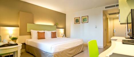 Quarto de hotel moderno com uma cama grande, uma escrivaninha, uma cadeira verde vibrante e um banheiro visível por uma porta aberta.