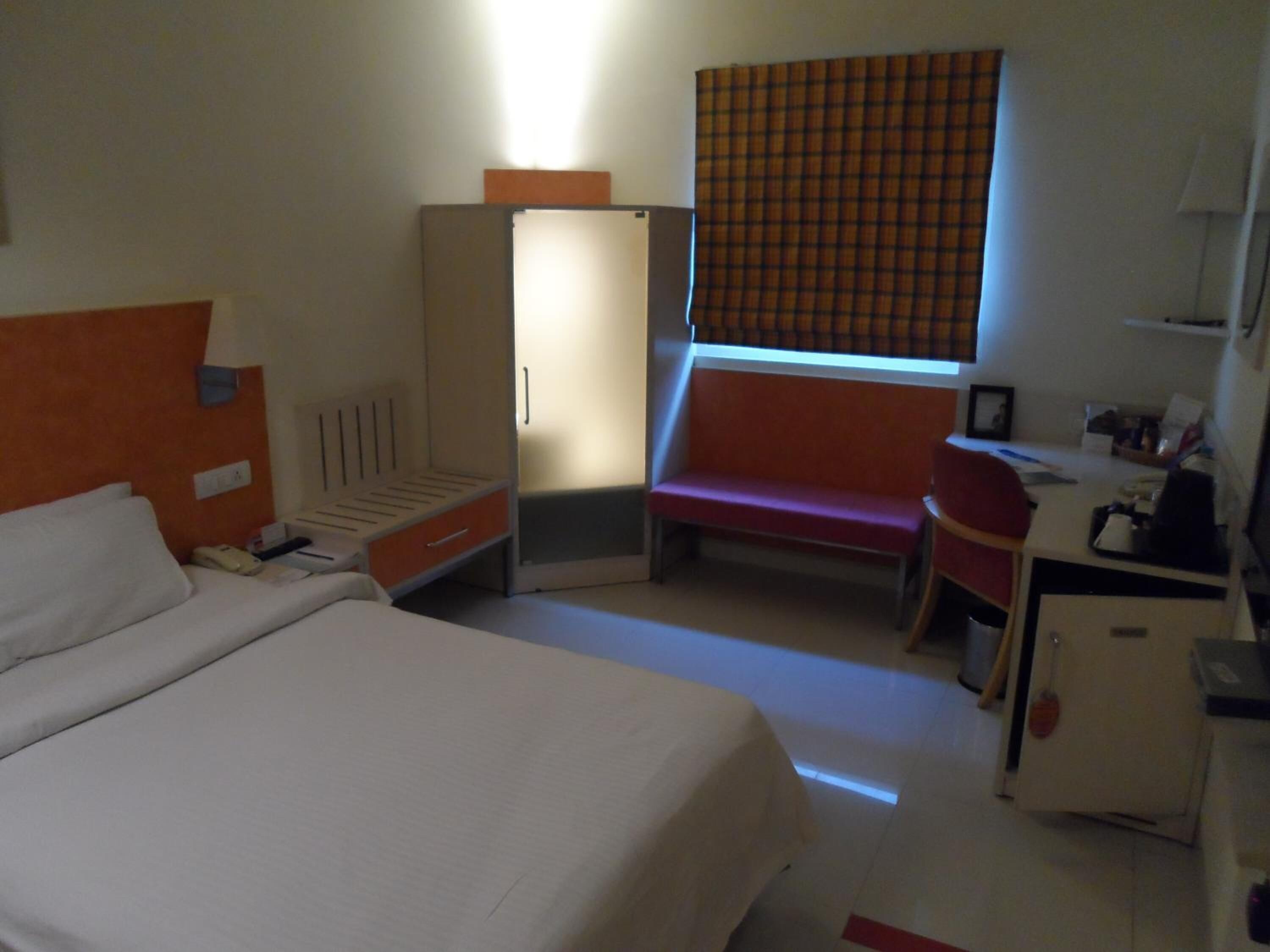 Quarto de hotel com cama, escrivaninha, cadeira, guarda-roupa e janela com persiana xadrez.