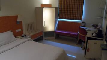 Quarto de hotel com cama, escrivaninha, cadeira, guarda-roupa e janela com persiana xadrez.