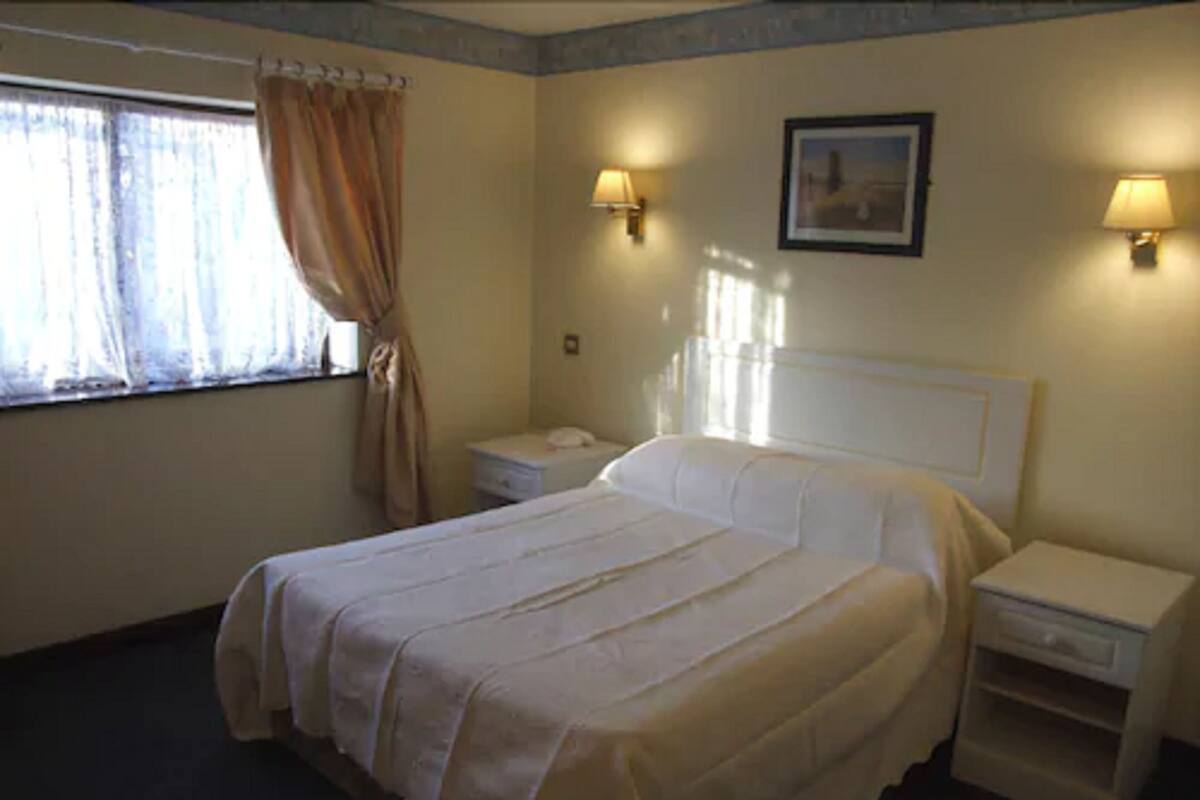 Quarto de hotel com cama, duas mesinhas de cabeceira, uma luminária, janela com cortinas e duas luminárias fixadas na parede.