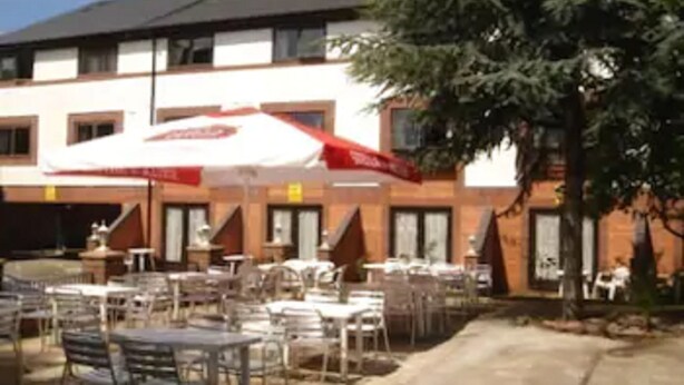 Uma área de café ao ar livre com mesas e cadeiras, um guarda-sol vermelho e um edifício com várias janelas.