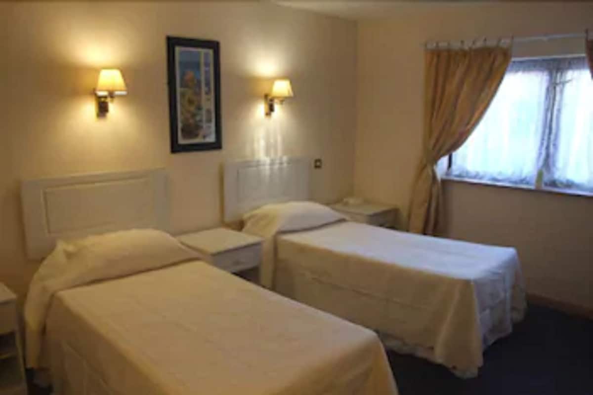 Quarto de hotel com duas camas de solteiro, uma mesa de cabeceira, uma luminária e uma janela com cortinas.