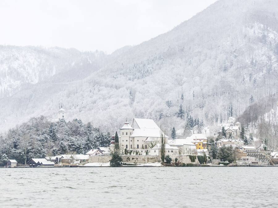 Um complexo de edifícios históricos à beira de um lago, cercado por árvores e colinas cobertas de neve.