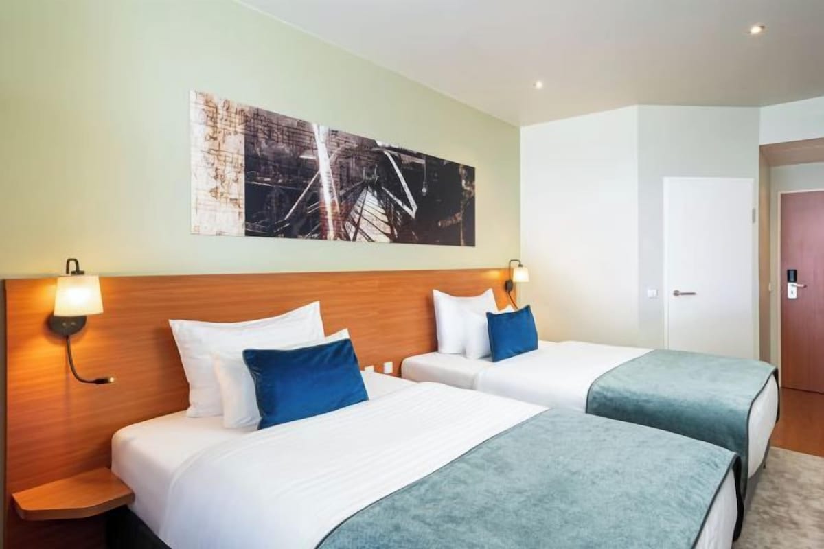Quarto de hotel com duas camas, cabeceira de madeira, obra de arte pendurada na parede e mesa de cabeceira com abajur.