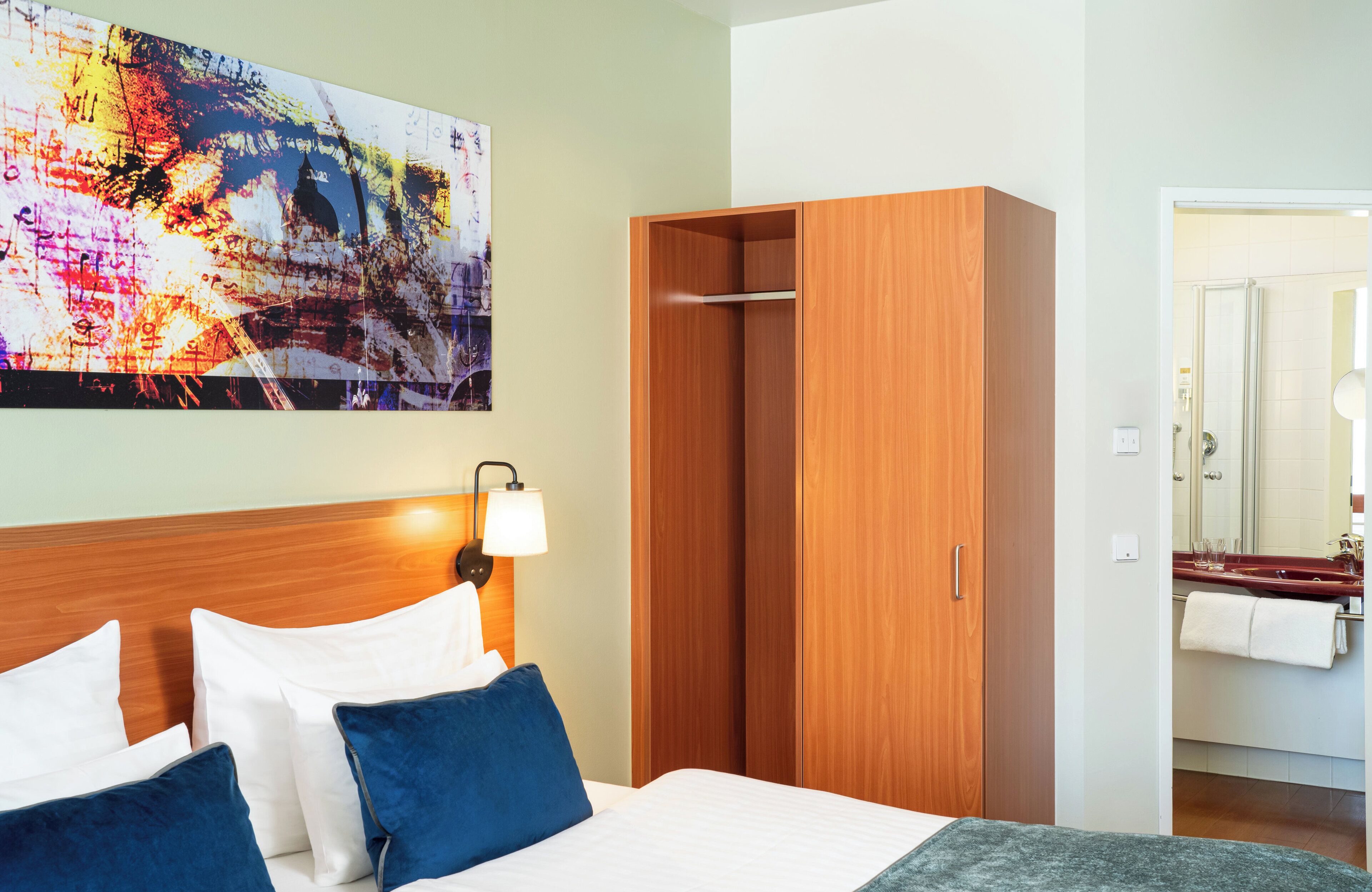 Quarto de hotel com uma cama, um guarda-roupa de madeira, uma mesa de cabeceira com abajur e uma pintura de paisagem urbana na parede.