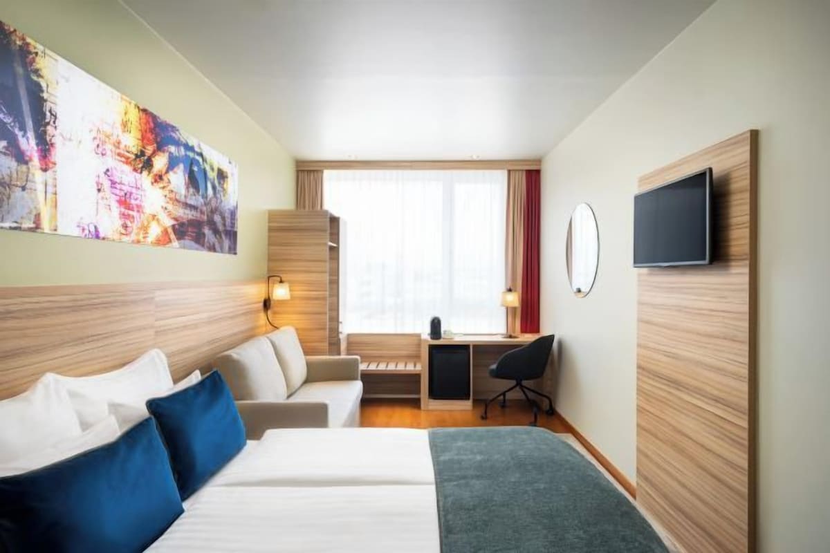 Quarto de hotel com cama, escrivaninha, cadeira, televisão e uma pintura grande na parede.