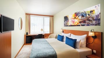 Quarto de hotel com uma cama grande, uma escrivaninha, uma cadeira, uma TV e um quadro na parede.
