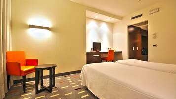 Quarto de hotel com uma cama, duas cadeiras laranja, uma mesa pequena, uma escrivaninha com cadeira e uma televisão.