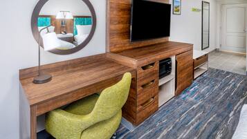 Quarto de hotel com escrivaninha de madeira, televisão de tela plana, espelho, cadeira verde e tapete estampado.