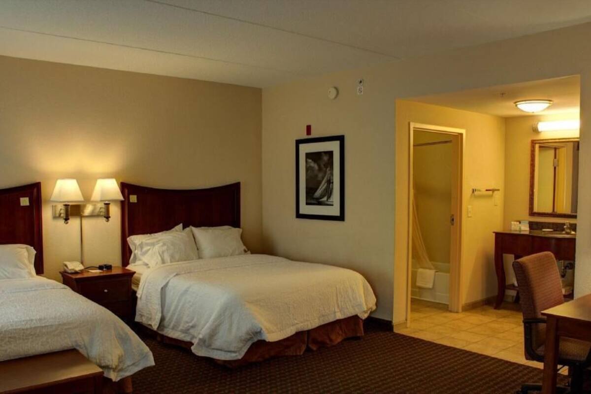 Quarto de hotel com duas camas, uma escrivaninha, uma cadeira, um espelho e um quadro na parede.