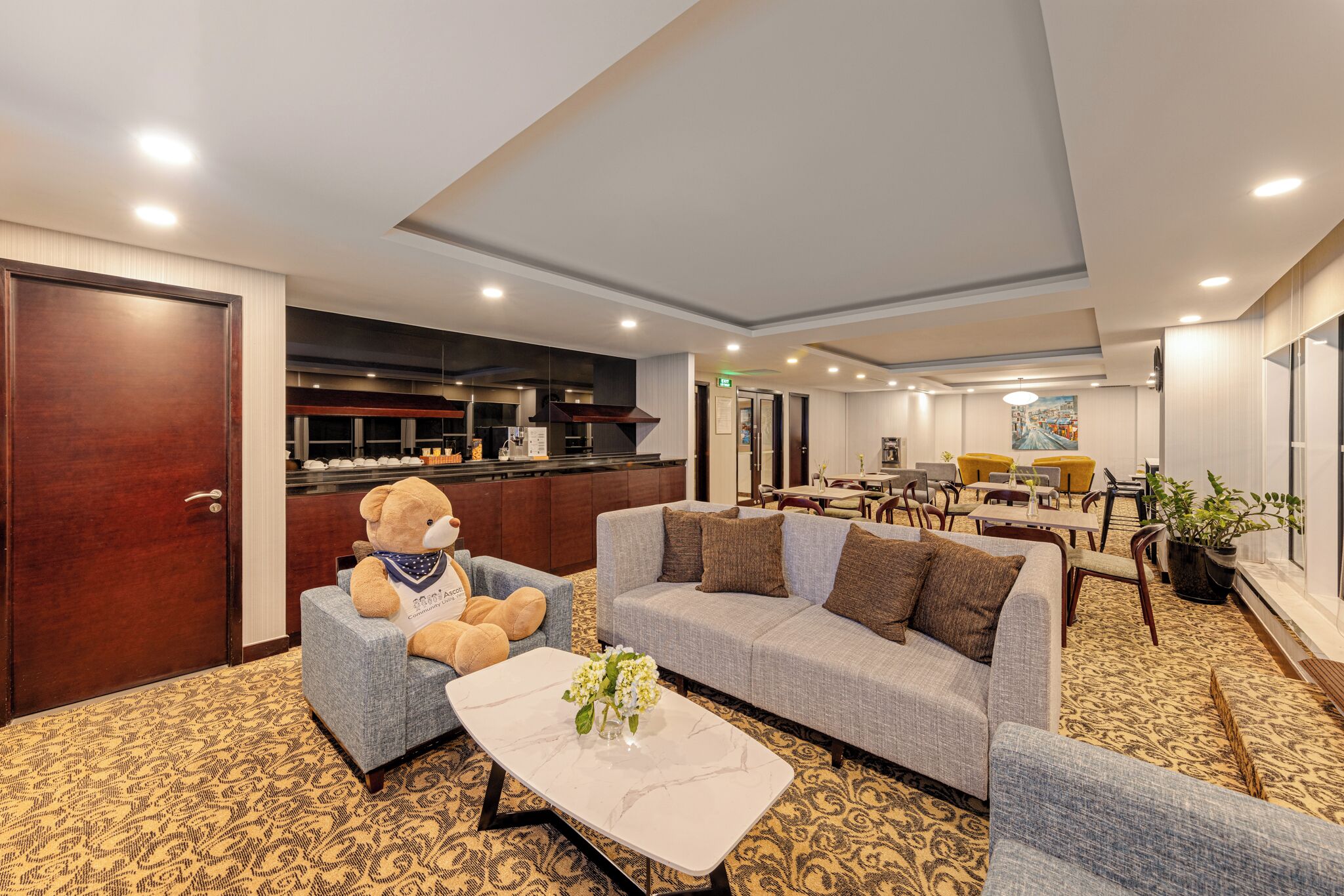 Um lobby de hotel com um sofá, uma poltrona, uma mesa de centro, um urso de pelúcia, uma receção e uma área de refeições.