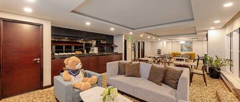 Um lobby de hotel com um sofá, uma poltrona, uma mesa de centro, um urso de pelúcia, uma receção e uma área de refeições.