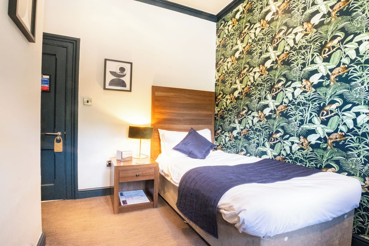 Quarto de hotel com cama, mesa de cabeceira e parede com padrão de folhagens.