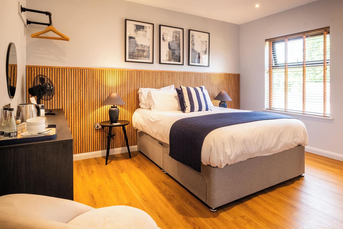 Quarto moderno com cabeceira em painel de madeira, uma cama grande com roupa de cama branca e azul, uma mesa de cabeceira com abajur e uma janela com persianas.