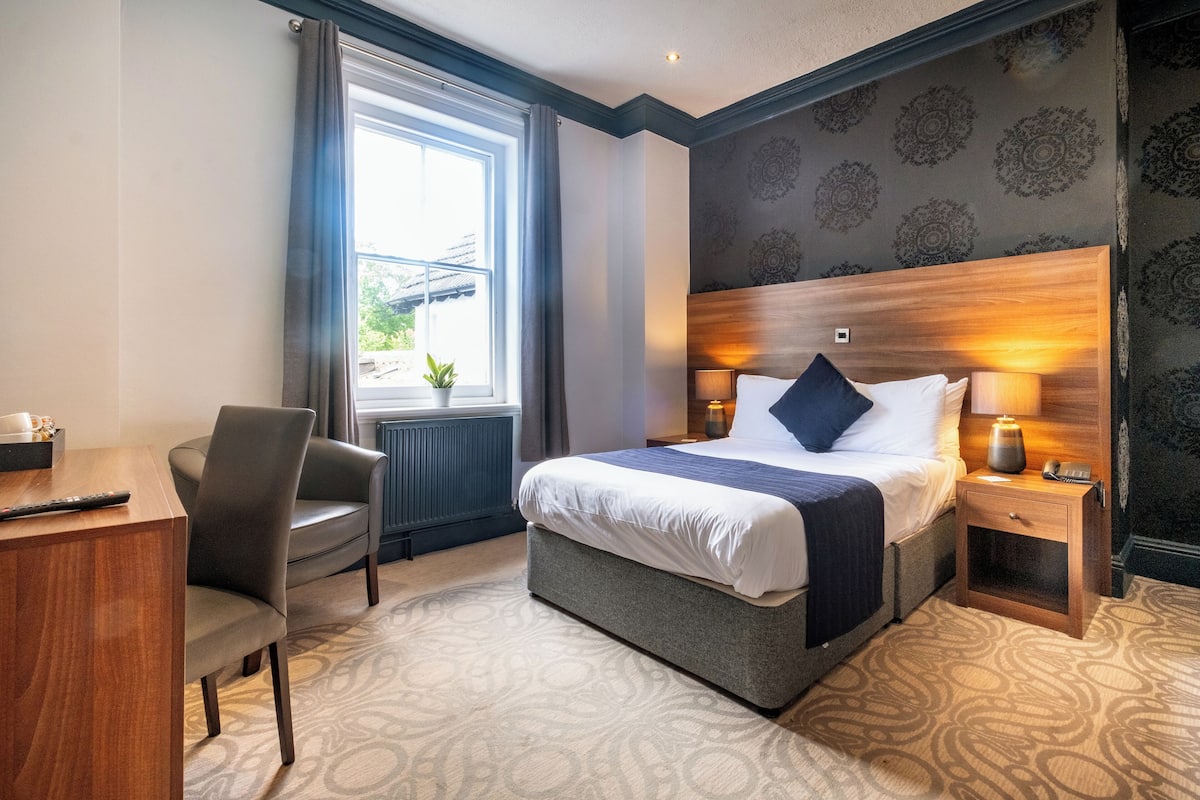 Quarto de hotel com uma cama grande, uma escrivaninha, uma cadeira, uma mesa de cabeceira e uma janela com cortinas.