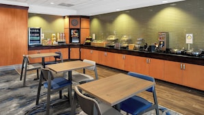 Espaço de café da manhã de hotel com mesas e cadeiras de madeira, um refrigerador e diversos displays de alimentos.