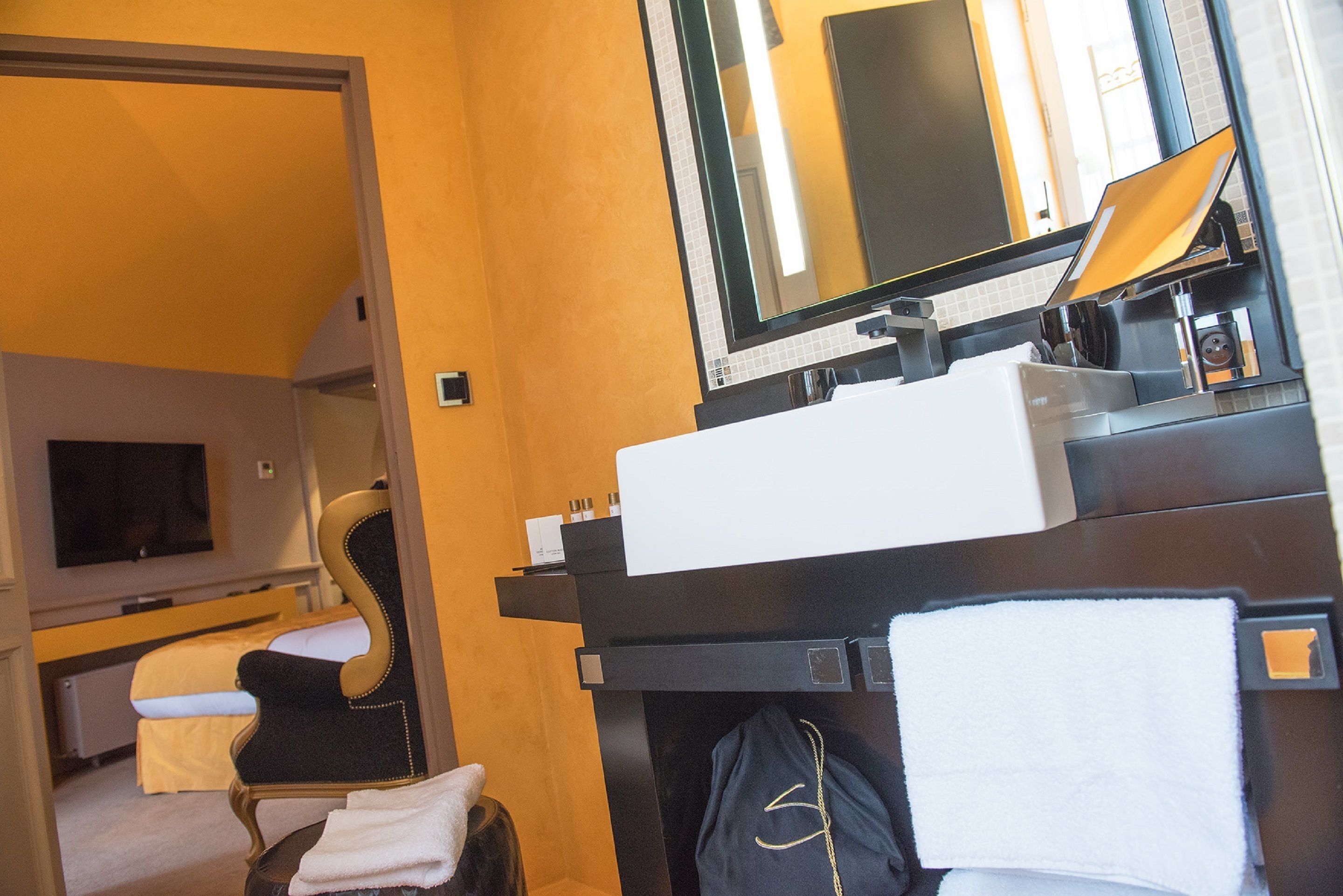 Quarto de hotel com cama, televisão, banheiro com pia e espelho, e porta-toalhas.