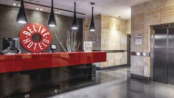 Uma área de recepção de hotel com um balcão vermelho, luminárias pendentes pretas, uma tela digital e uma placa com a inscrição "BELIEVE HOTEL".