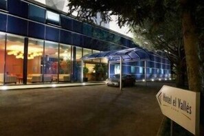 Exterior de hotel à noite com fachada de vidro, área externa coberta e placa com o nome do hotel.
