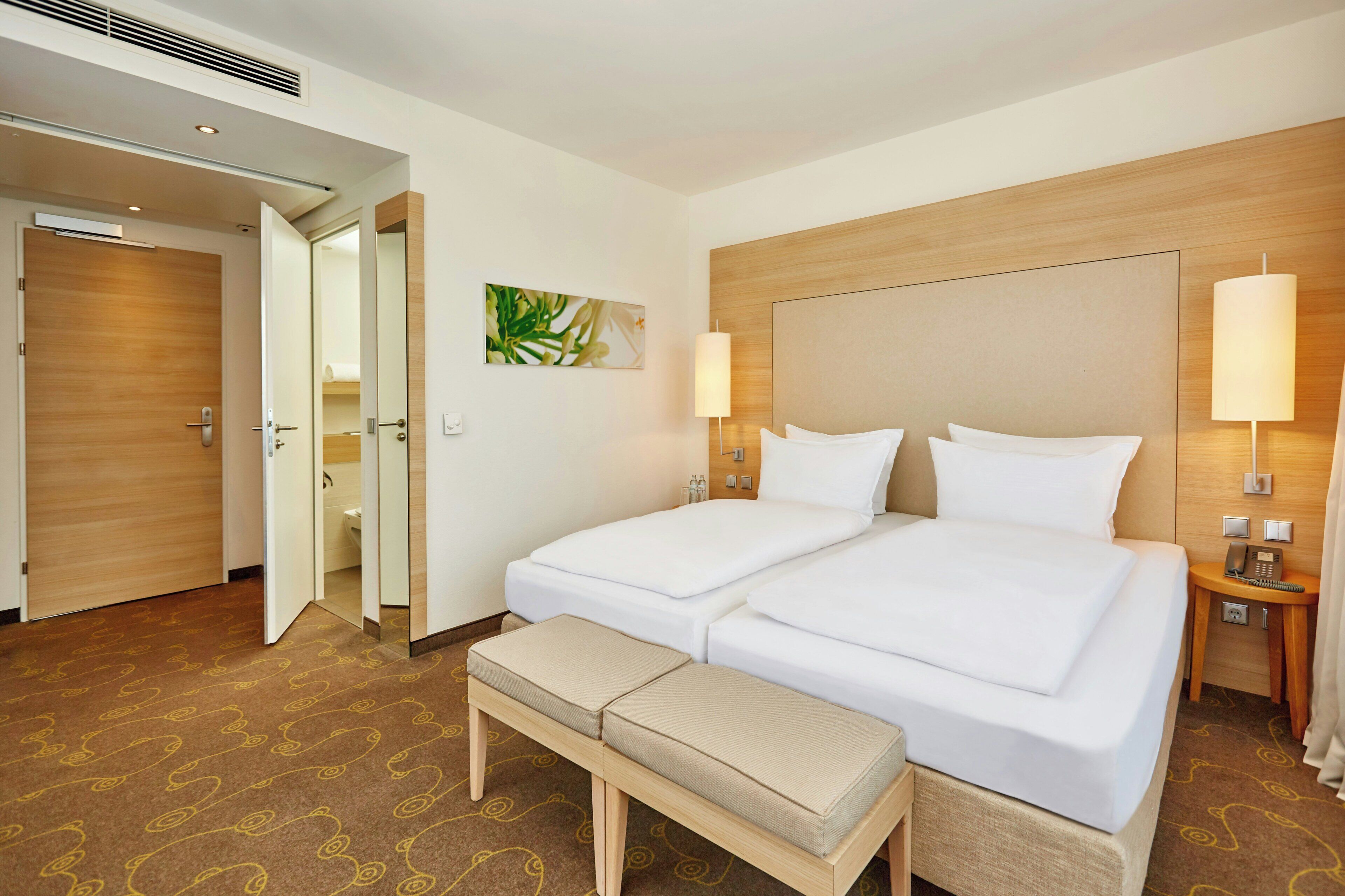 Quarto de hotel com duas camas, um banco, uma mesinha de cabeceira com telefone e uma luminária na parede.