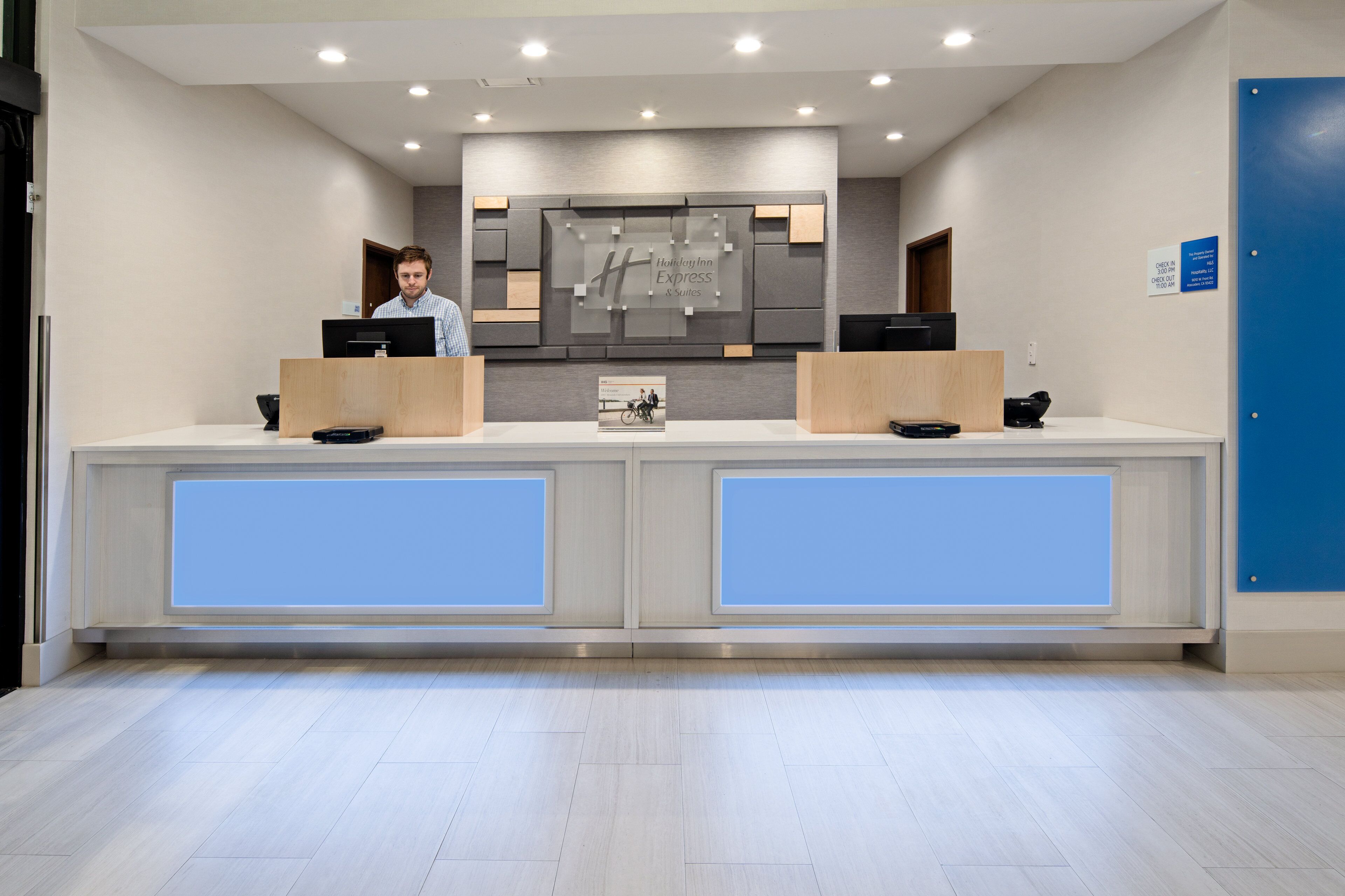 Uma receção com dois postos de computador, um painel de visualização grande na parede e uma porta azul.