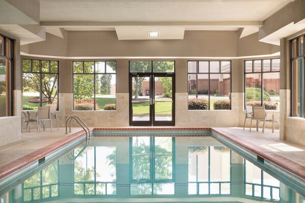 Piscina com paredes de vidro transparente, rodeada por paredes bege e grandes janelas com vista para um jardim ajardinado.