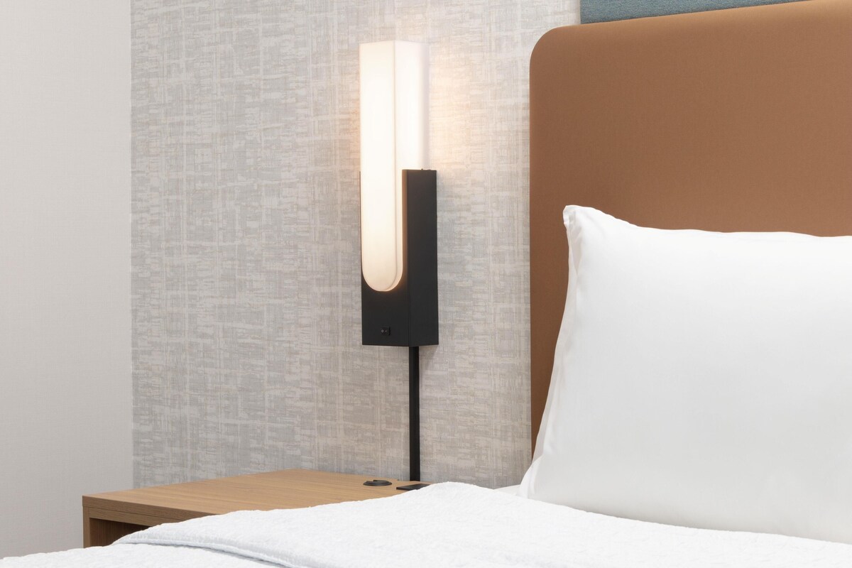 Uma cama com travesseiro branco e uma mesa de cabeceira com luminária moderna.